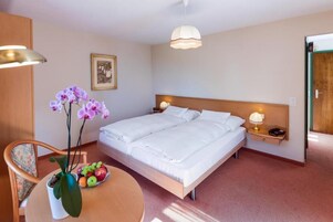 Quarto de hotel com cama de casal, mesas de cabeceira com luminárias, uma cadeira, uma mesa redonda com frutas e uma orquídea em vaso, e um quadro na parede.