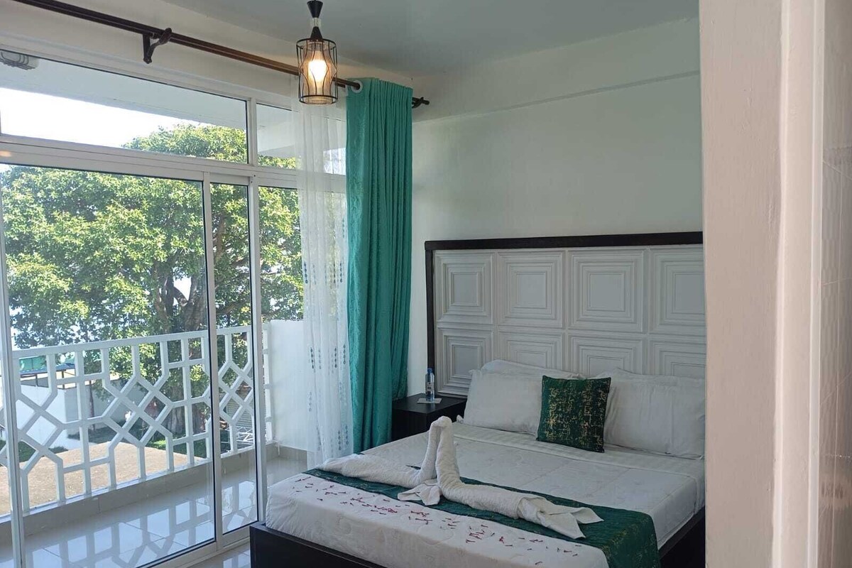 Quarto com cama, mesa de cabeceira, janela com cortinas e vista para o verde lá fora.