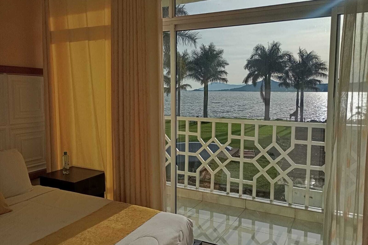 Quarto de hotel com uma janela grande, cama, mesa de cabeceira e vista para o mar e palmeiras.