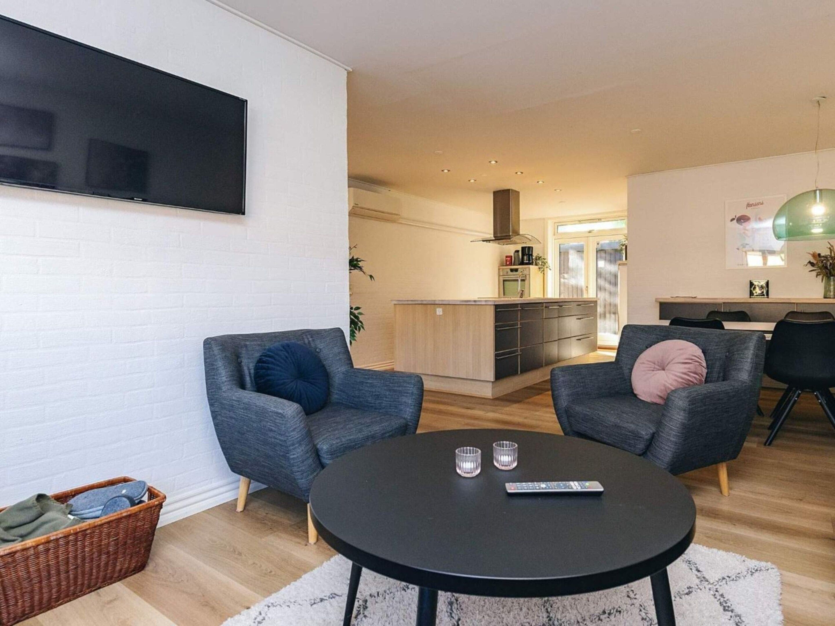 Uma sala de estar moderna com dois poltronas cinzas, uma mesa de centro redonda preta e uma TV de tela plana fixada na parede.