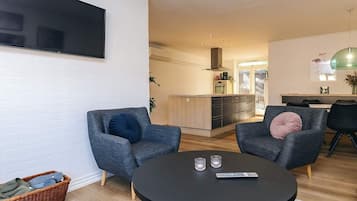Uma sala de estar moderna com dois poltronas cinzas, uma mesa de centro redonda preta e uma TV de tela plana fixada na parede.
