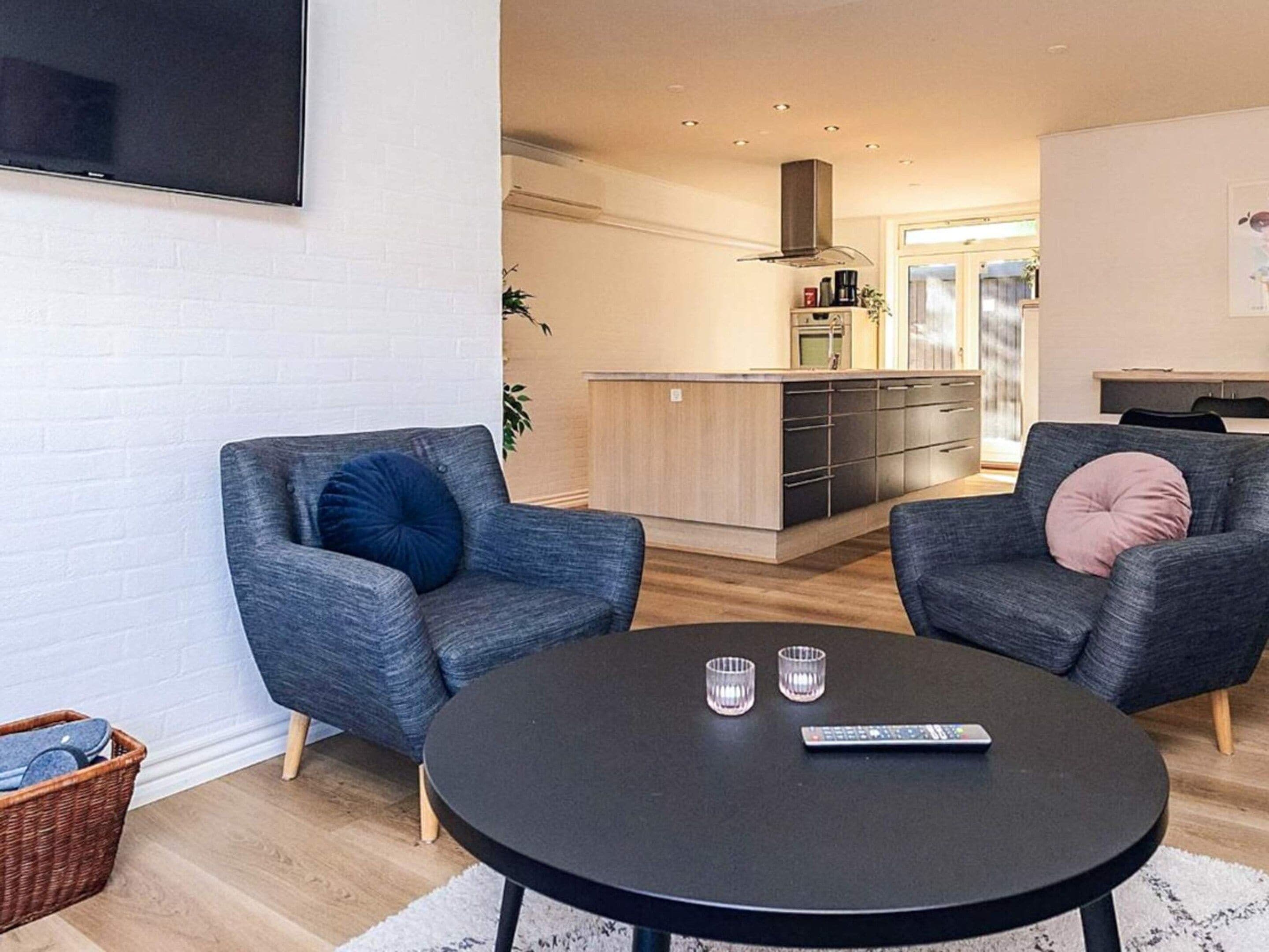 Uma sala de estar moderna com duas poltronas cinzentas, uma mesa de centro redonda preta e uma televisão fixada em uma parede de tijolos brancos.