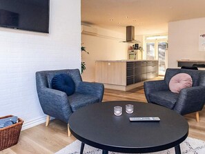 Uma sala de estar moderna com duas poltronas cinzentas, uma mesa de centro redonda preta e uma televisão fixada em uma parede de tijolos brancos.