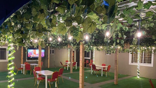 Uma área de refeições ao ar livre com mesas e cadeiras, rodeada por vegetação ornamental e luzes de corda.