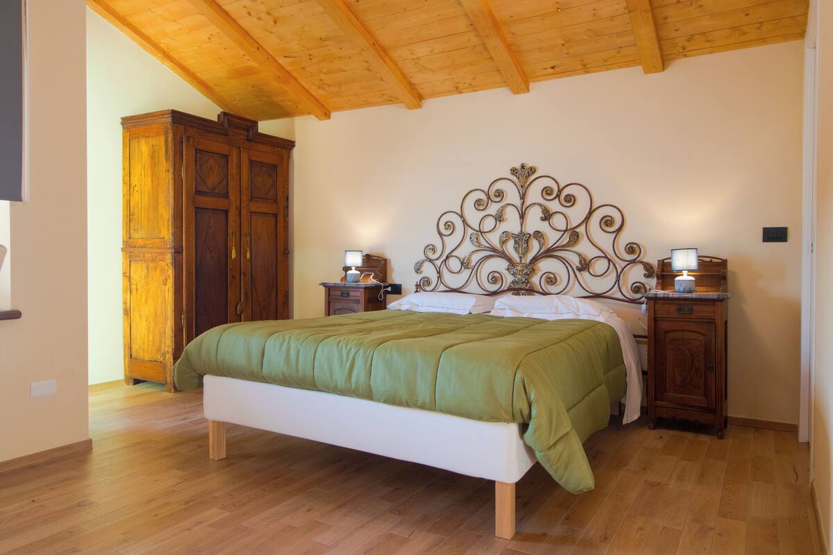 Quarto com cama de madeira, mesa de cabeceira com luminária, guarda-roupa e um objeto decorativo na parede.