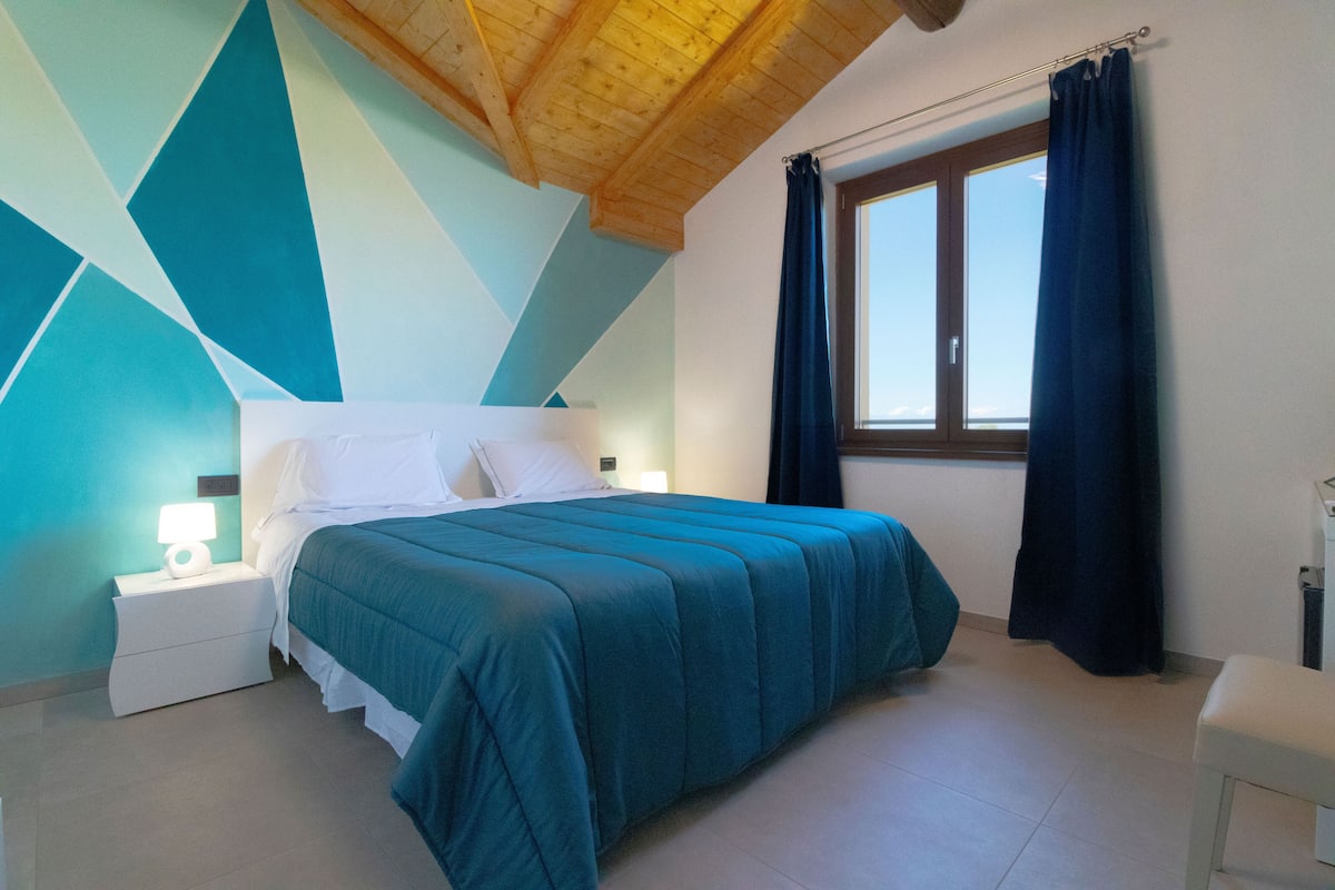 Quarto com cama, mesa de cabeceira e janela com cortinas.