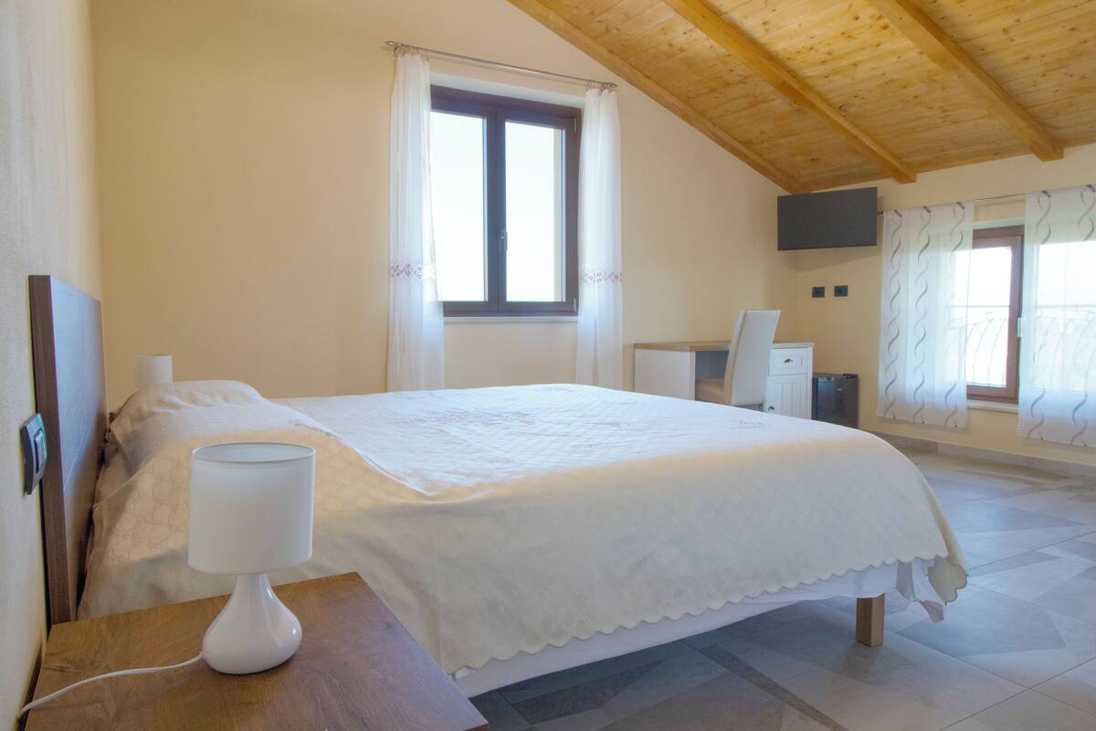 Quarto com uma cama, luminária de cabeceira, janela, televisão e uma pequena escrivaninha.