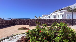 Um hotel à beira-mar com parede de pedra, jardim com flores vermelhas e céu azul límpido.