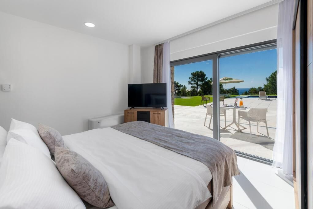 Quarto com cama, TV sobre suporte de madeira e varanda com mobiliário externo.