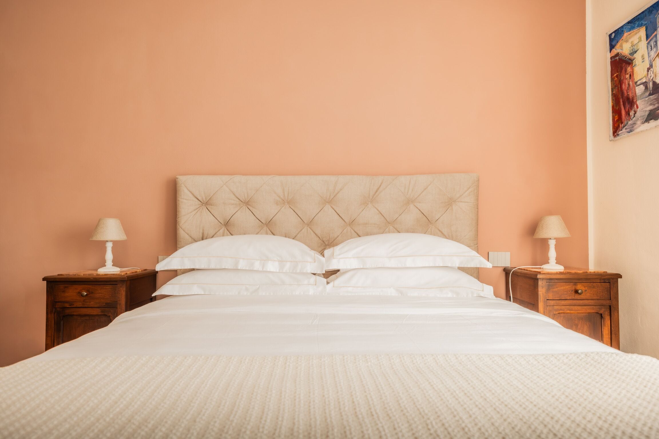 Uma cama com cabeceira estofada, duas mesas de cabeceira com abajures e um quadro na parede.