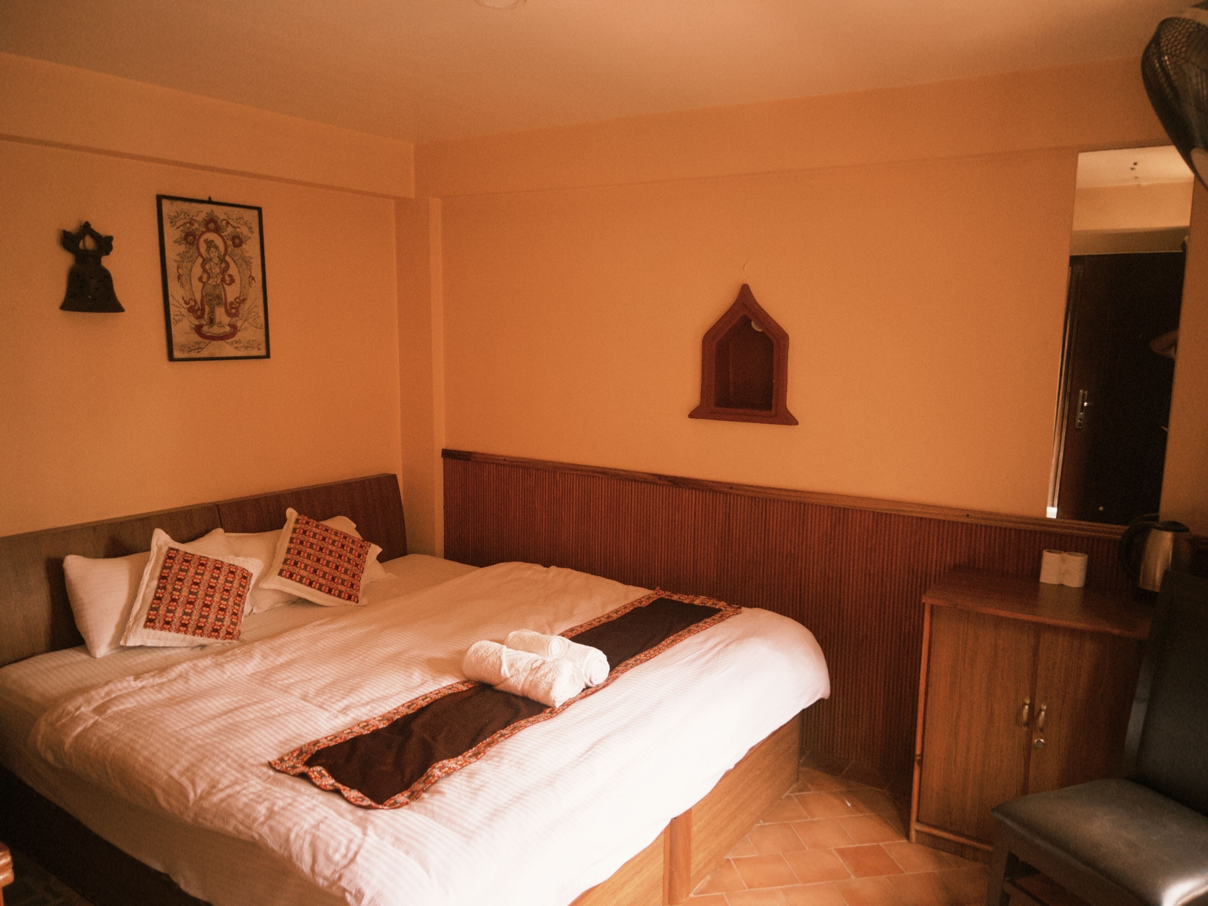Cama bem arrumada com colcha estampada e travesseiros, cabeceira de madeira, um pequeno espelho decorativo e um armário de madeira com uma cadeira.
