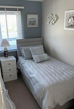 Quarto com cama, mesa de cabeceira, abajur e janela com persianas.