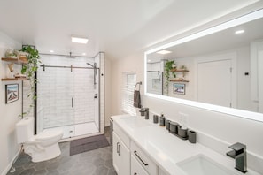 Uma casa de banho moderna com cabine de duche em vidro, sanita branca, espelho grande e toucador branco com lavatório.