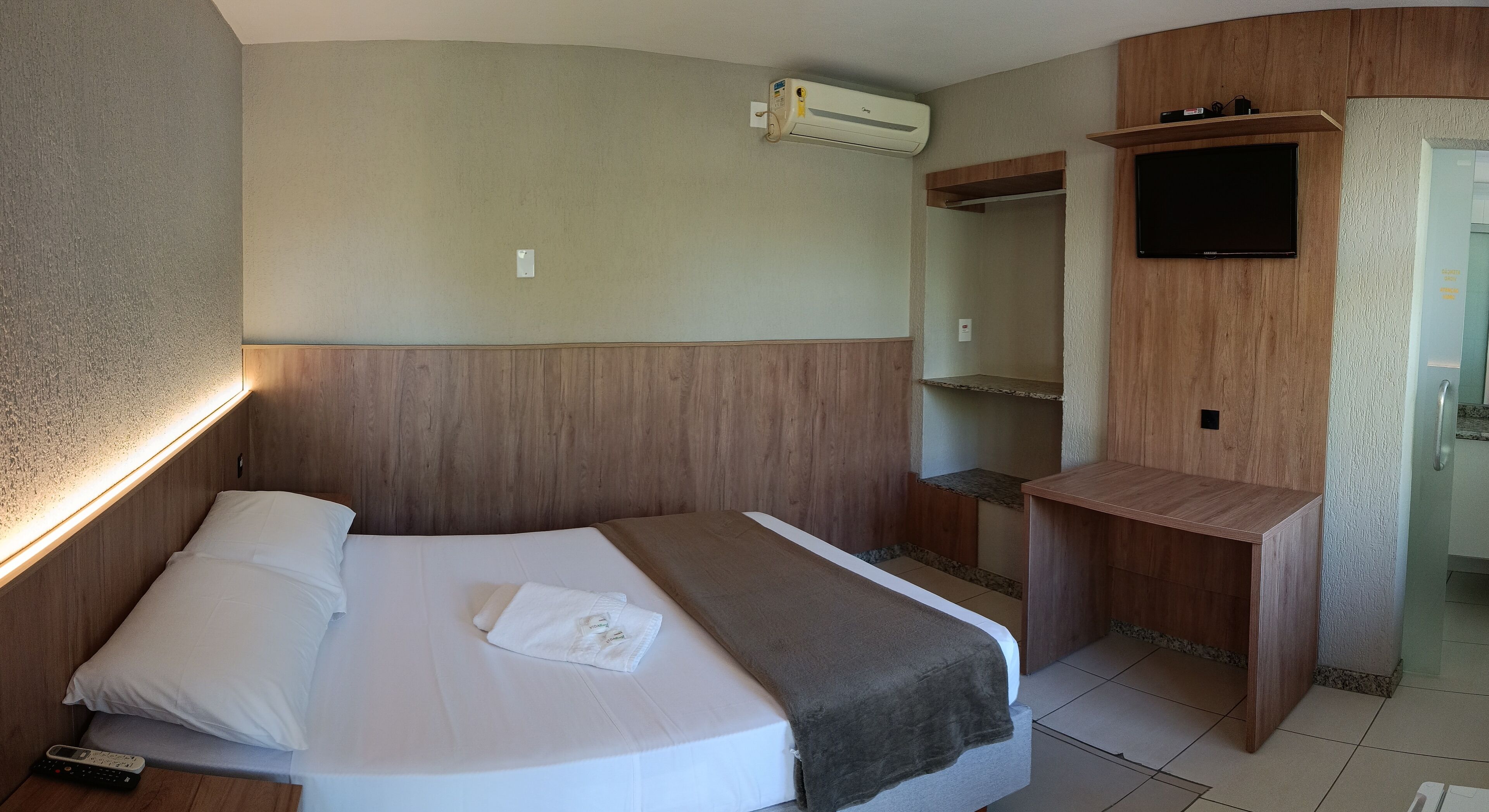 Quarto de hotel com cama, mesa de cabeceira e televisão integrada.