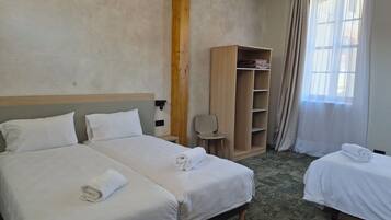 Quarto de hotel com duas camas, um guarda-roupa, uma cadeira e janela com cortinas.