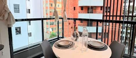 Uma área de refeições moderna com uma mesa redonda, cadeiras pretas e vista para um edifício de tijolos vermelhos através de grandes janelas.