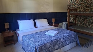 Uma cama com colcha estampada, duas mesas de cabeceira com luminárias, uma cama alta à direita e uma parede padronizada.