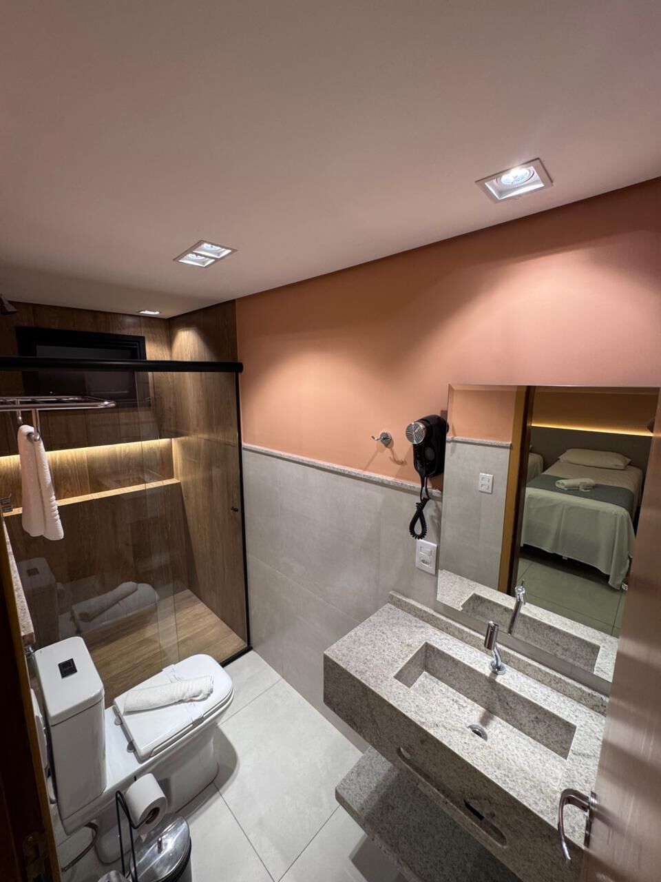 Uma casa de banho moderna com cabine de duche em vidro, sanita, lavatório com espelho e secador de cabelo fixado na parede.