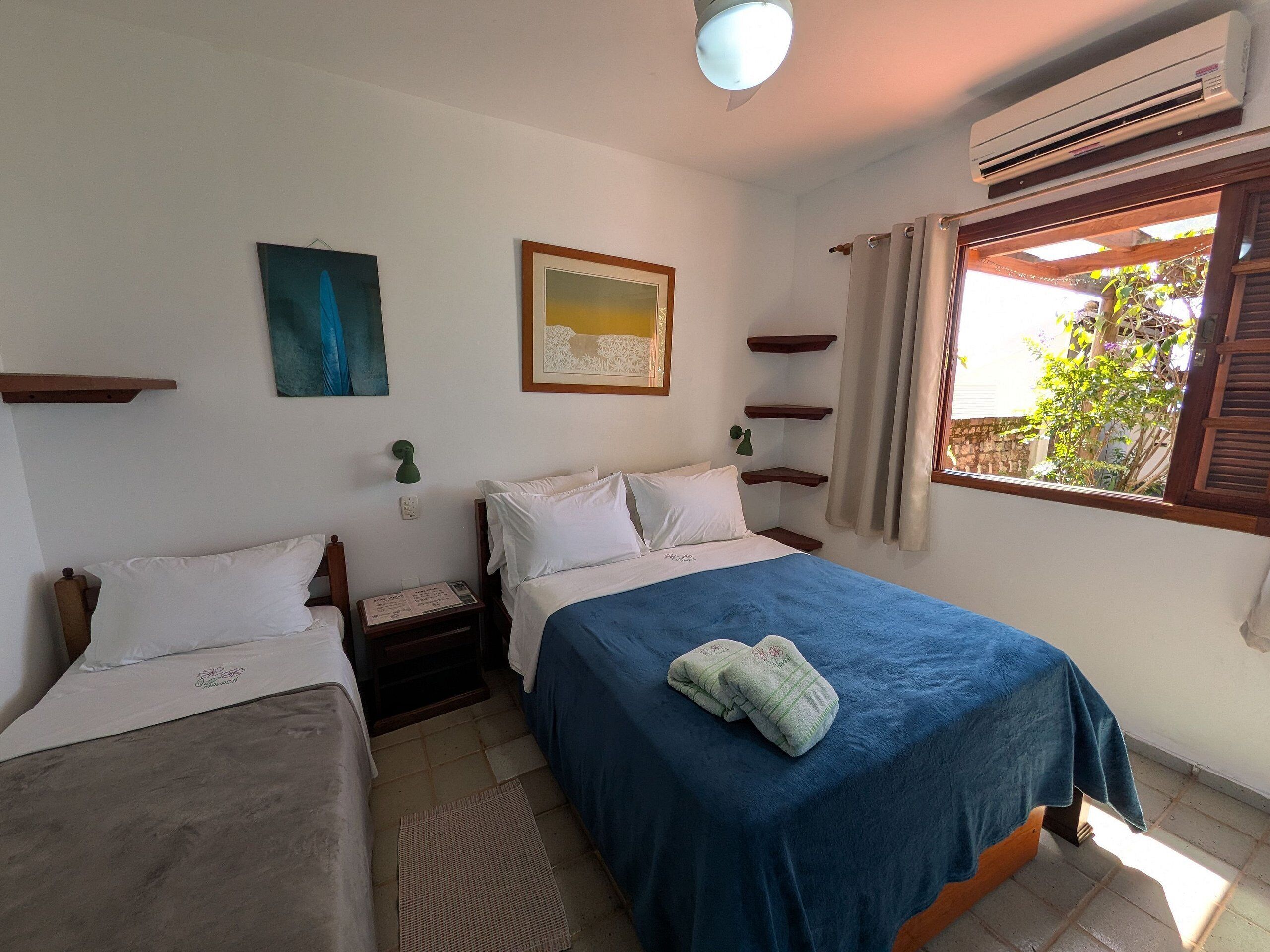 Quarto com duas camas, janela com cortinas, ar condicionado fixado na parede e uma pequena mesa com abajur.