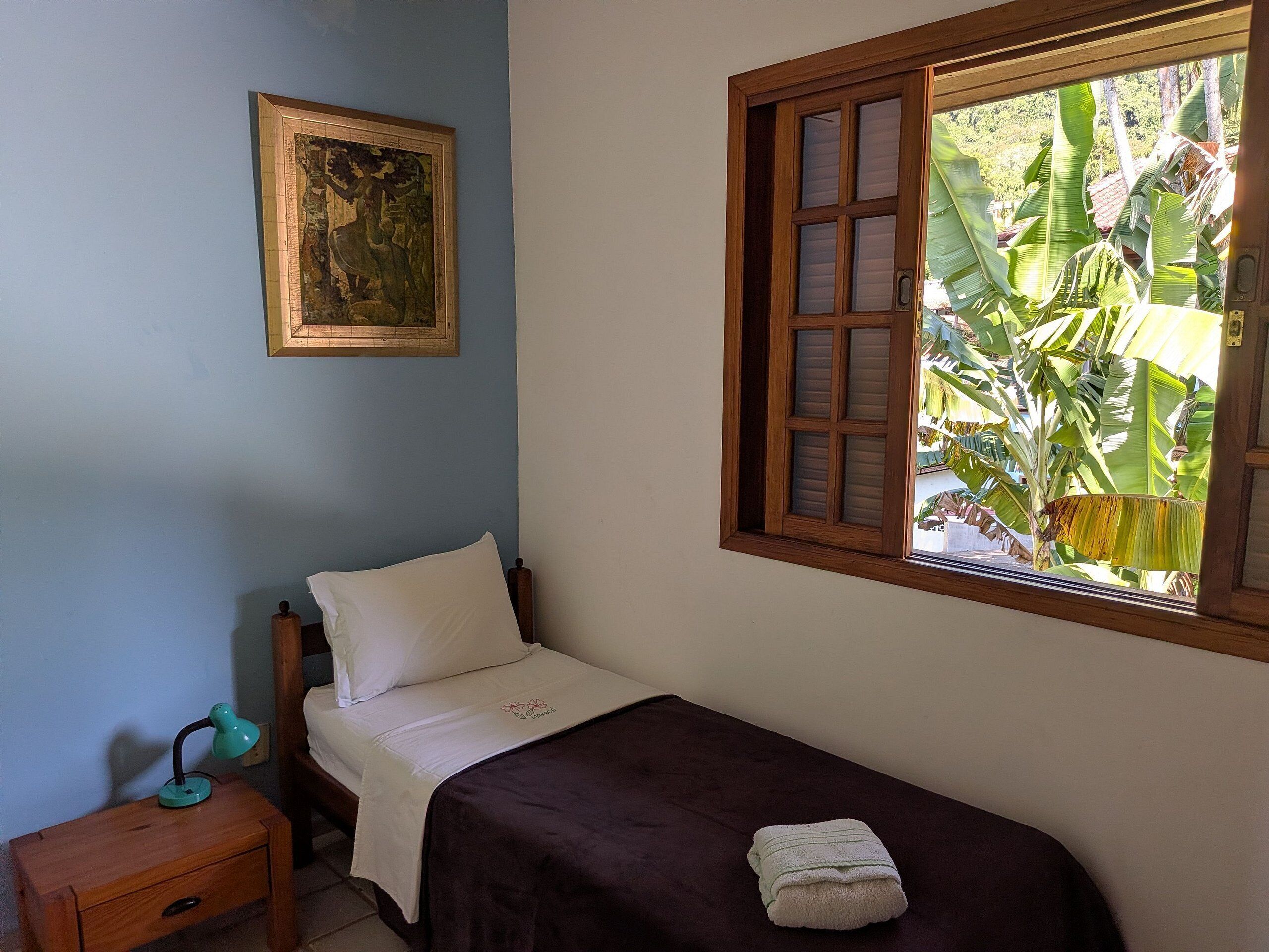 Uma cama de solteiro com um cobre-leito escuro, um travesseiro branco, uma pequena mesa de cabeceira com abajur, uma janela com vista para bananeiras e um quadro na parede.