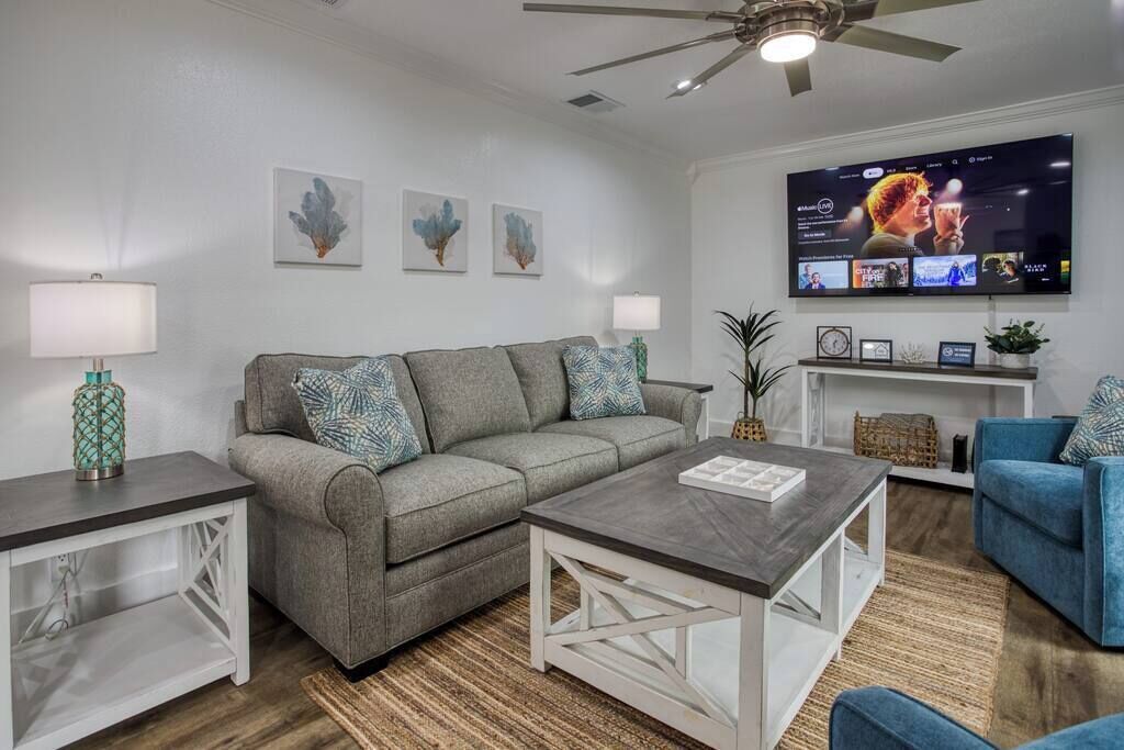 Uma sala de estar com um sofá cinzento, uma mesa de centro, uma TV fixada na parede e um ventilador de teto.