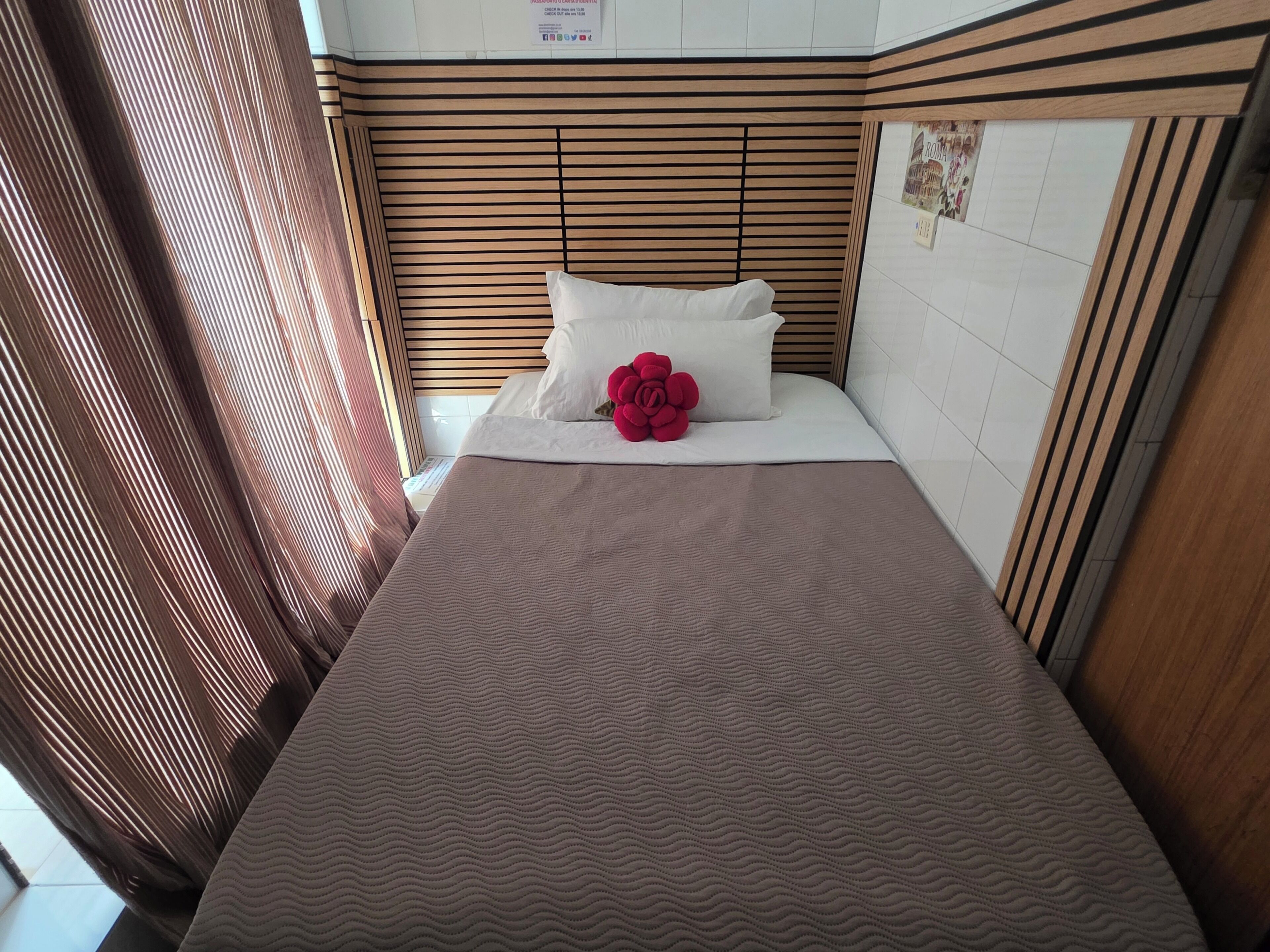 Quarto de hotel com uma cama, cabeceira de madeira, parede branca com um cartaz e uma janela com persianas.
