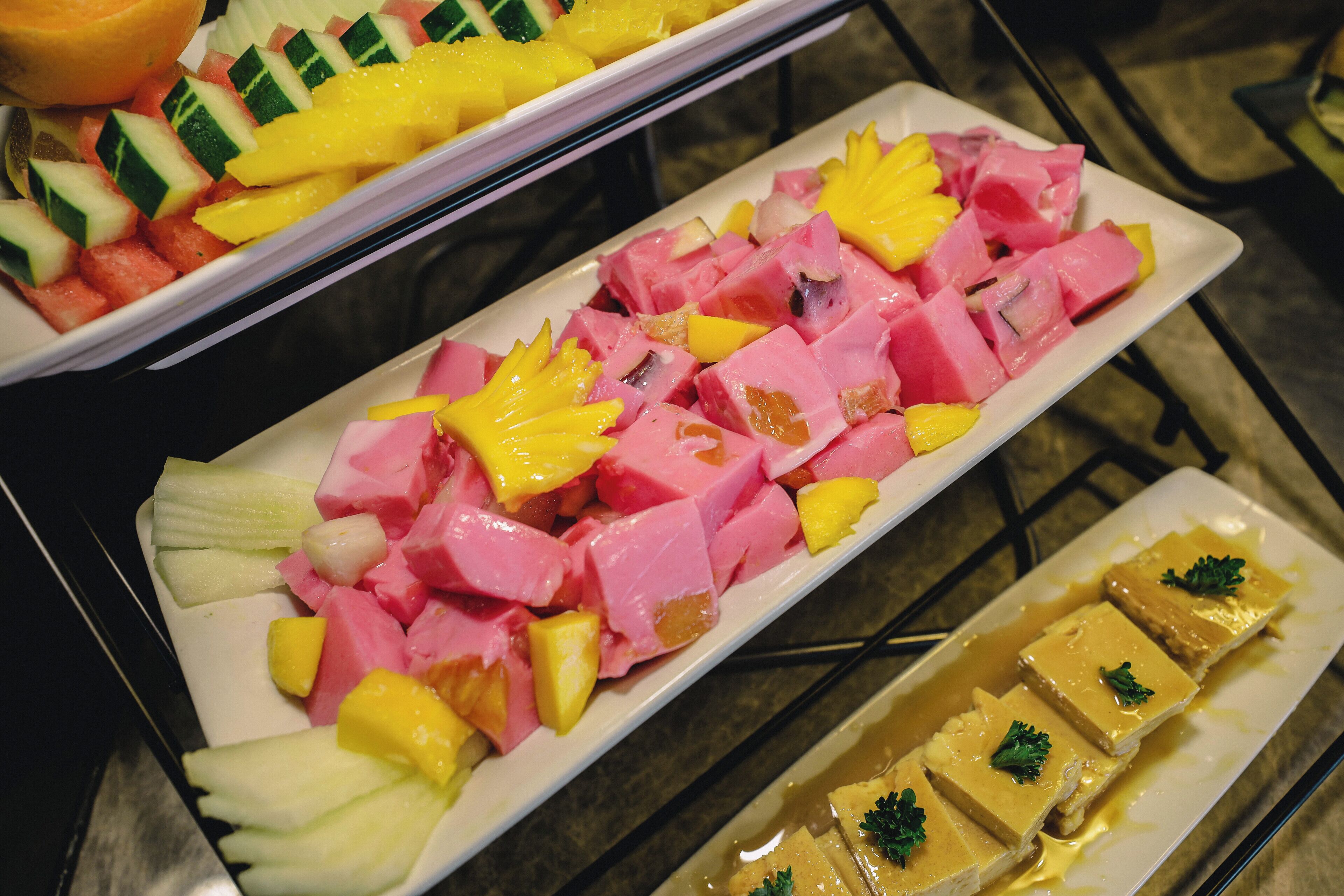 Buffet com diversos alimentos, incluindo cubos rosa e amarelos, possivelmente frutas, e um prato com molho marrom.