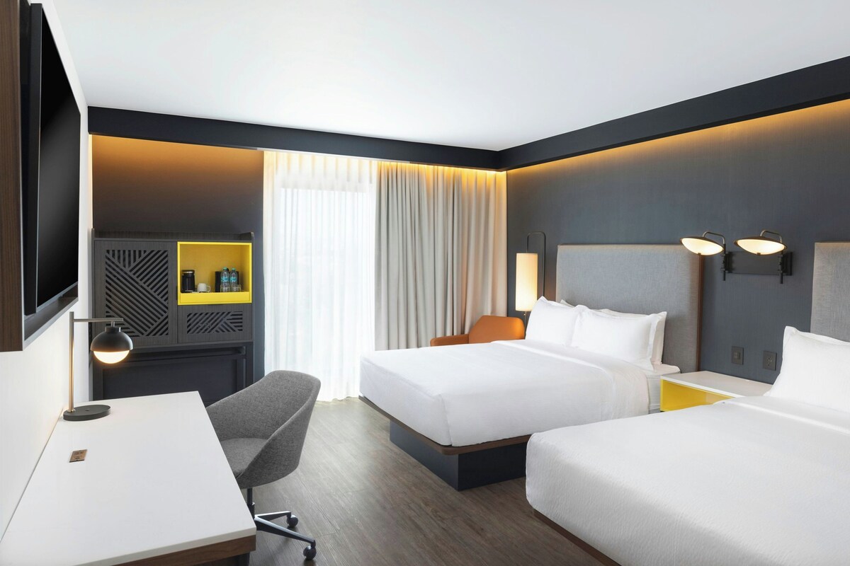 Quarto de hotel com cama, mesas de cabeceira, escrivaninha, cadeira, televisão e janela com cortinas.