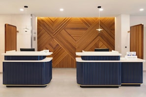 Uma área de recepção de hotel com dois balcões, uma parede de madeira com padrão geométrico e iluminação embutida no teto.