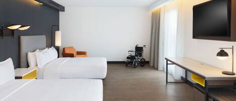 Quarto de hotel com duas camas, uma escrivaninha, uma televisão e uma cadeira de rodas.