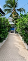 Uma passarela pavimentada leva a um edifício com porta azul, cercado por vegetação exuberante e uma palmeira.