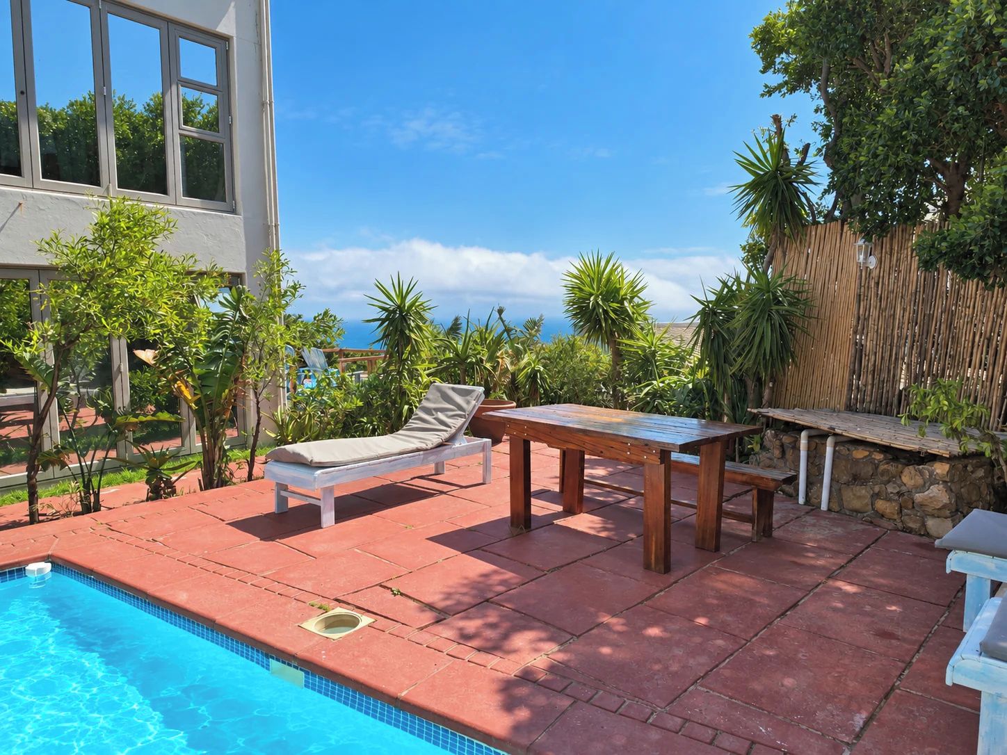 Área da piscina com espreguiçadeira, mesa de madeira e vista para o oceano.