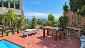 Área da piscina com espreguiçadeira, mesa de madeira e vista para o oceano.