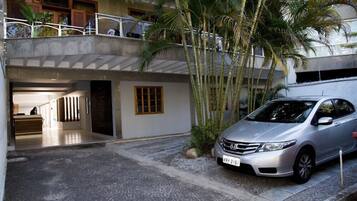 Um edifício moderno com um carro estacionado em frente, uma varanda com plantas e uma entrada visível.