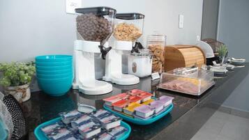 Uma mesa de buffet com distribuidores de comida, bandejas com alimentos embalados e um recipiente de madeira para comida.