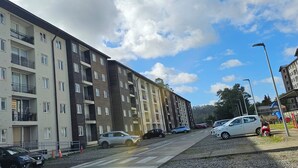 Um edifício residencial com vários andares, varandas, área de estacionamento com carros e um parquinho ao fundo.