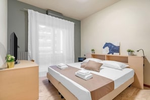 Quarto de hotel com cama, escrivaninha, radiador, janela com cortinas, televisão presa à parede e uma pequena planta.