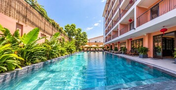 Piscina com espreguiçadeiras e guarda-sóis, cercada por vegetação exuberante e um edifício com varandas.
