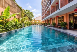 Piscina com espreguiçadeiras e guarda-sóis, rodeada por vegetação e um edifício com varandas.