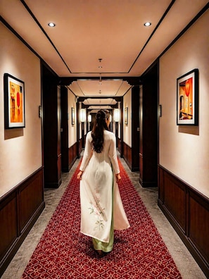 Um corredor de hotel com carpete vermelho, obras de arte emolduradas nas paredes e uma pessoa caminhando pelo corredor.