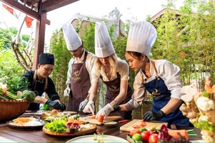 Um grupo de chefs prepara alimentos ao ar livre, com diversos vegetais e uma mesa de madeira.