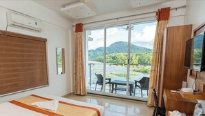 Quarto de hotel com cama, televisão, escrivaninha e vista para um rio e montanhas.
