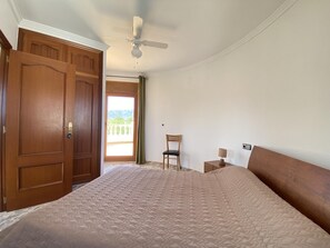Quarto com cama, cadeira, mesa de cabeceira, ventoinha no teto e vista através de uma porta.