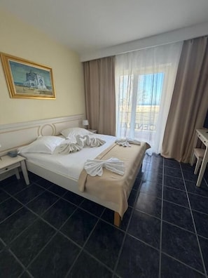 Quarto de hotel com cama, mesa de cabeceira, escrivaninha e janela com cortinas.
