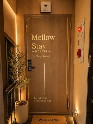 Entrada de hotel com uma placa indicando 'Mellow Stay - HOSTEL' e 'CFTV EM FUNCIONAMENTO'.