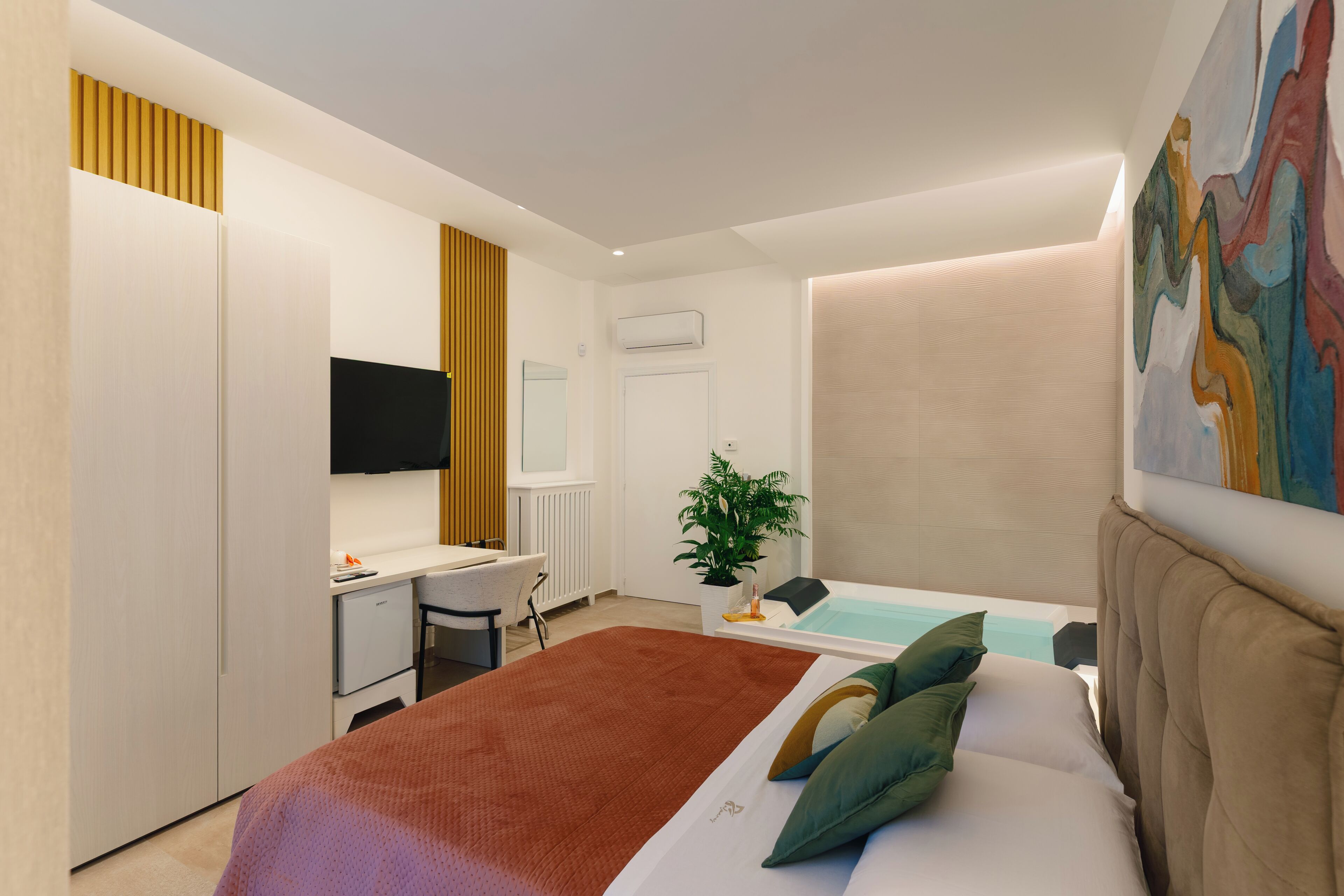 Quarto de hotel com cama, escrivaninha, cadeira, mesinha, planta e um quadro na parede.