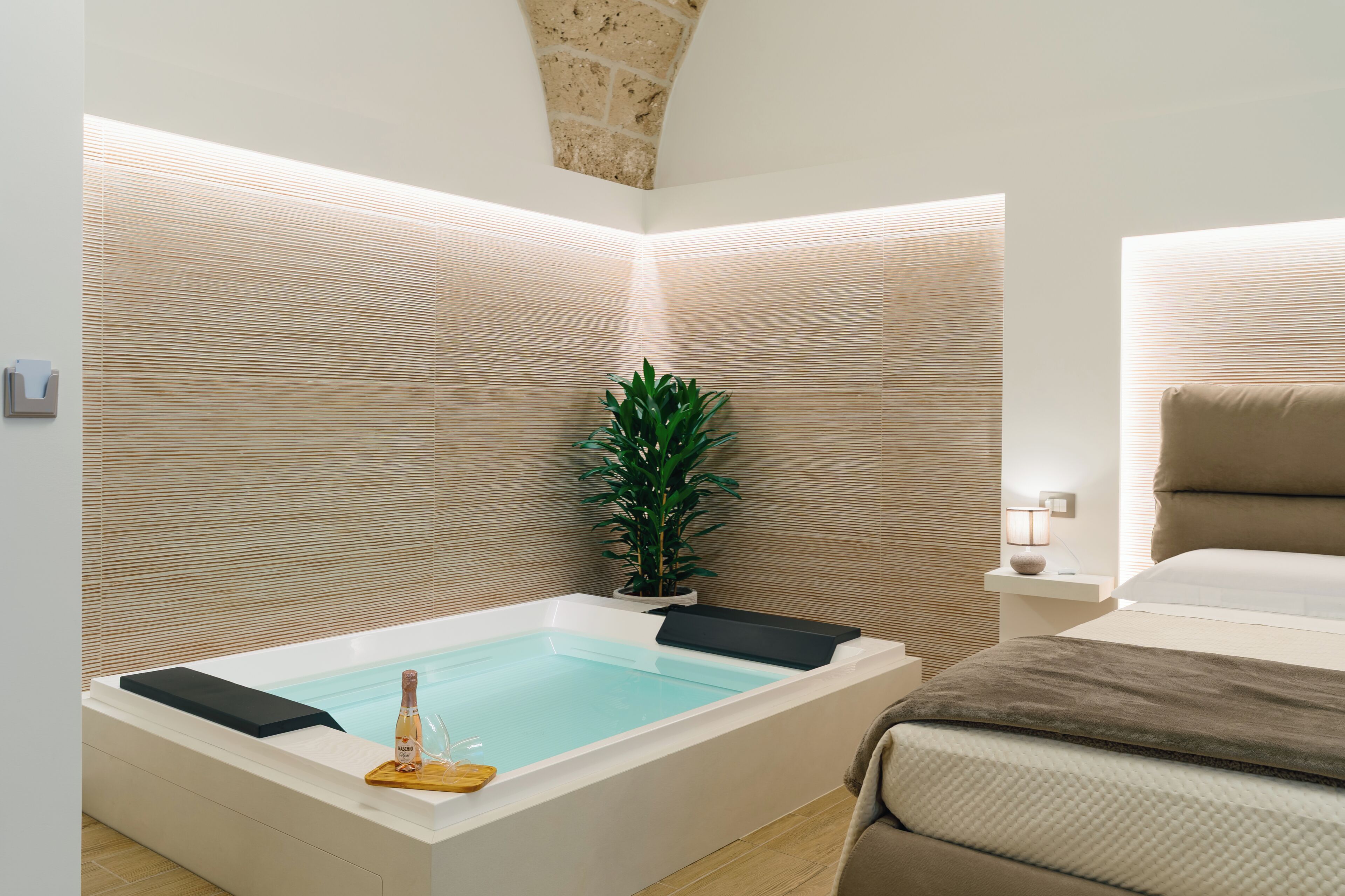 Um spa moderno com uma banheira de hidromassagem retangular, uma planta em vaso, uma mesinha de cabeceira com abajur e uma cama com cabeceira marrom.