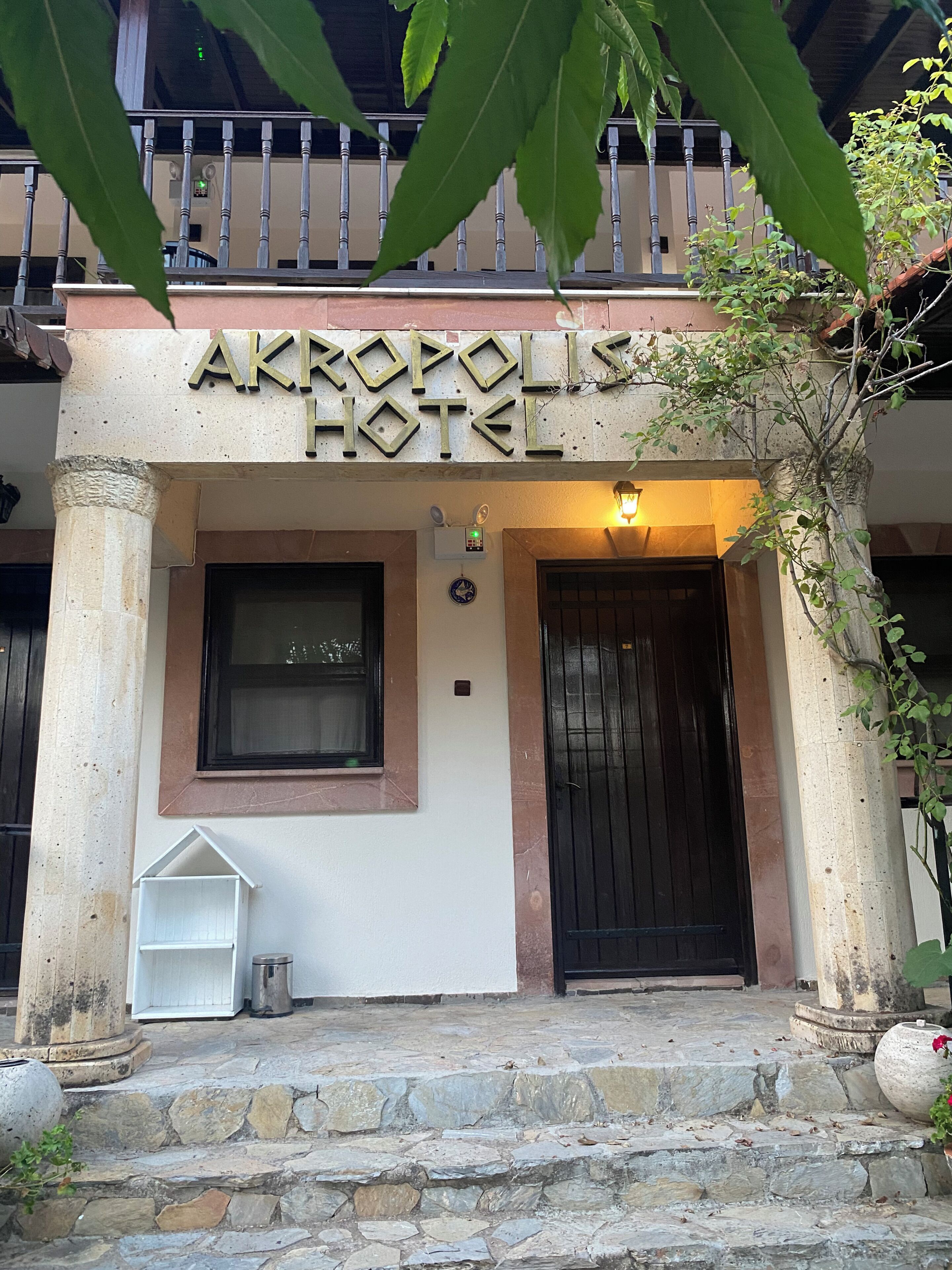 Entrada de hotel com uma placa indicando 'AKROPOLIS HOTEL', uma porta preta, uma janela e uma pequena construção em forma de casa branca.