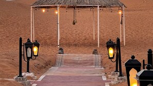 Uma trilha no deserto que leva a uma construção com telhado de palha, com luzes e lanternas penduradas em ambos os lados.