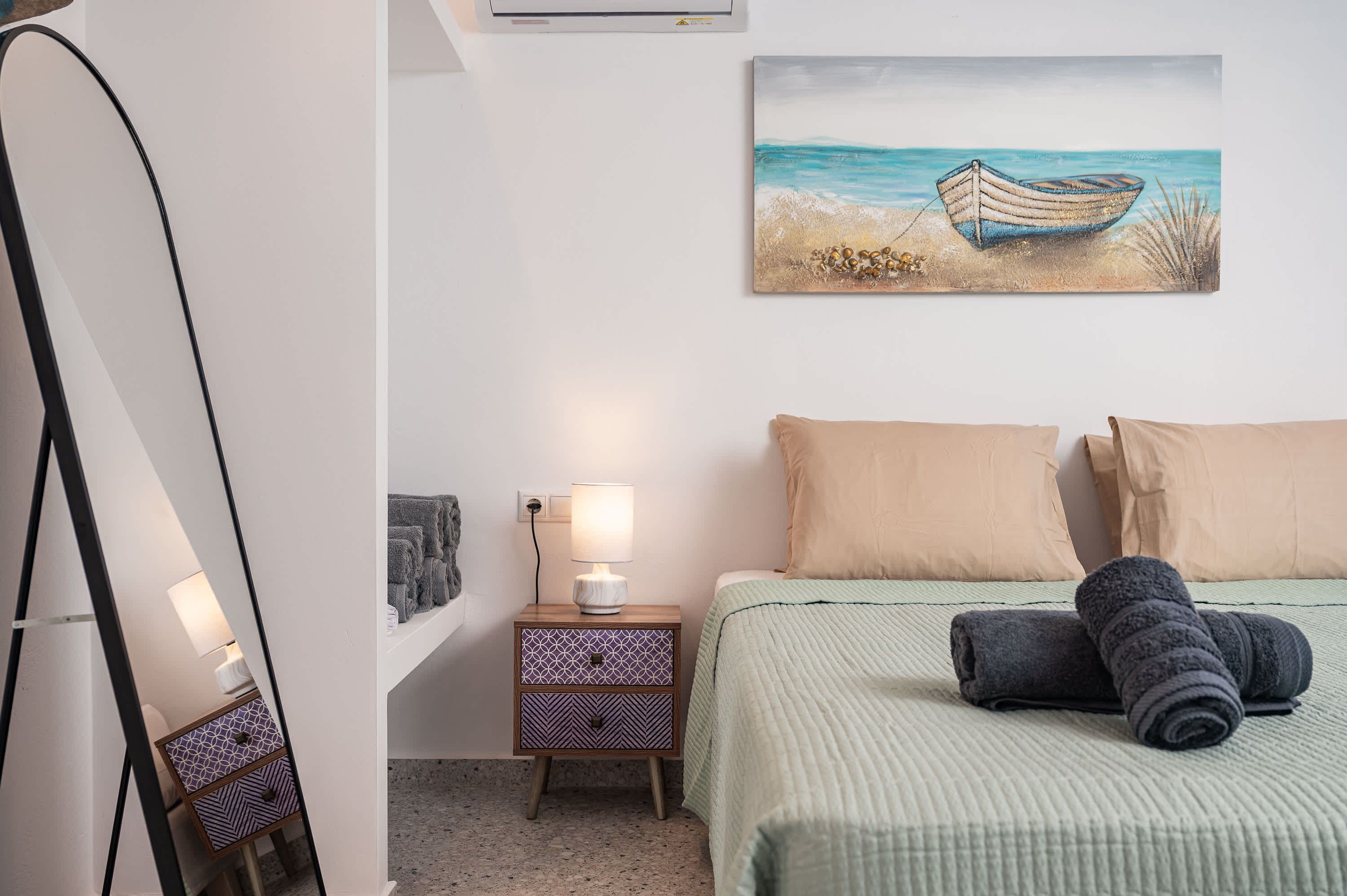 Quarto com cama, travesseiros, mesa de cabeceira com abajur, quadro de um barco na parede e espelho.